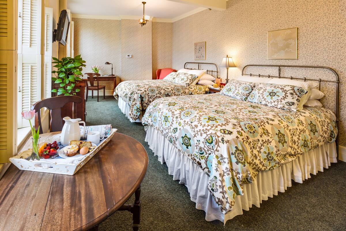 Quarto de hotel com duas camas, uma escrivaninha, uma cadeira e uma mesa de jantar com itens de café da manhã.