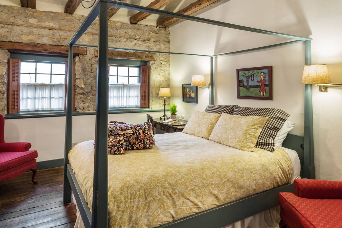Uma cama de dossel com colcha florida, uma poltrona vermelha, uma mesinha de cabeceira com abajur e um quadro na parede.