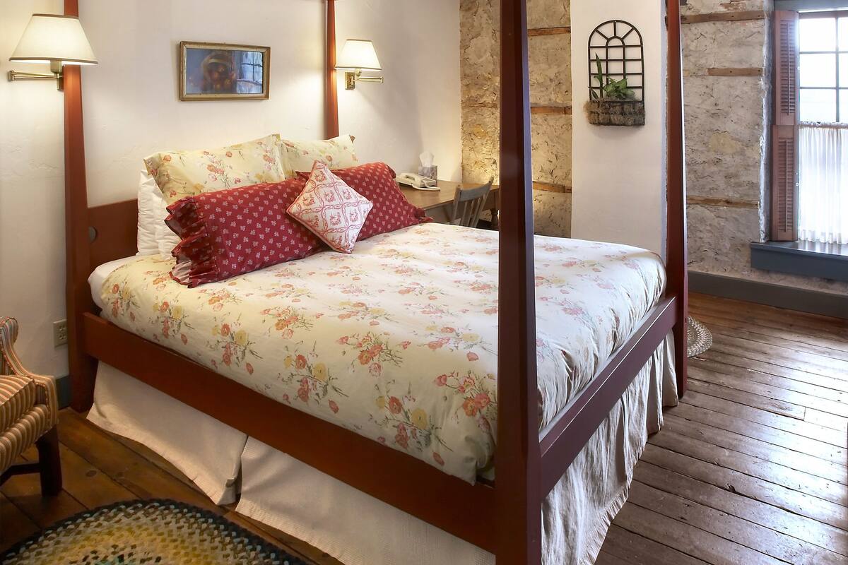 Uma cama com dossel e roupa de cama florida, piso de madeira, parede de pedra e janela com cortina.