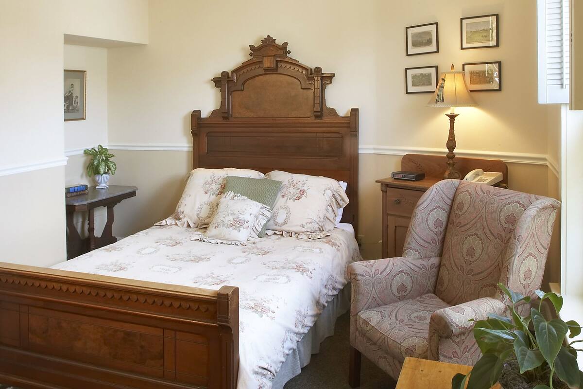 Um quarto com uma cama de madeira, uma cadeira, uma mesa de cabeceira, um abajur e quadros nas paredes.