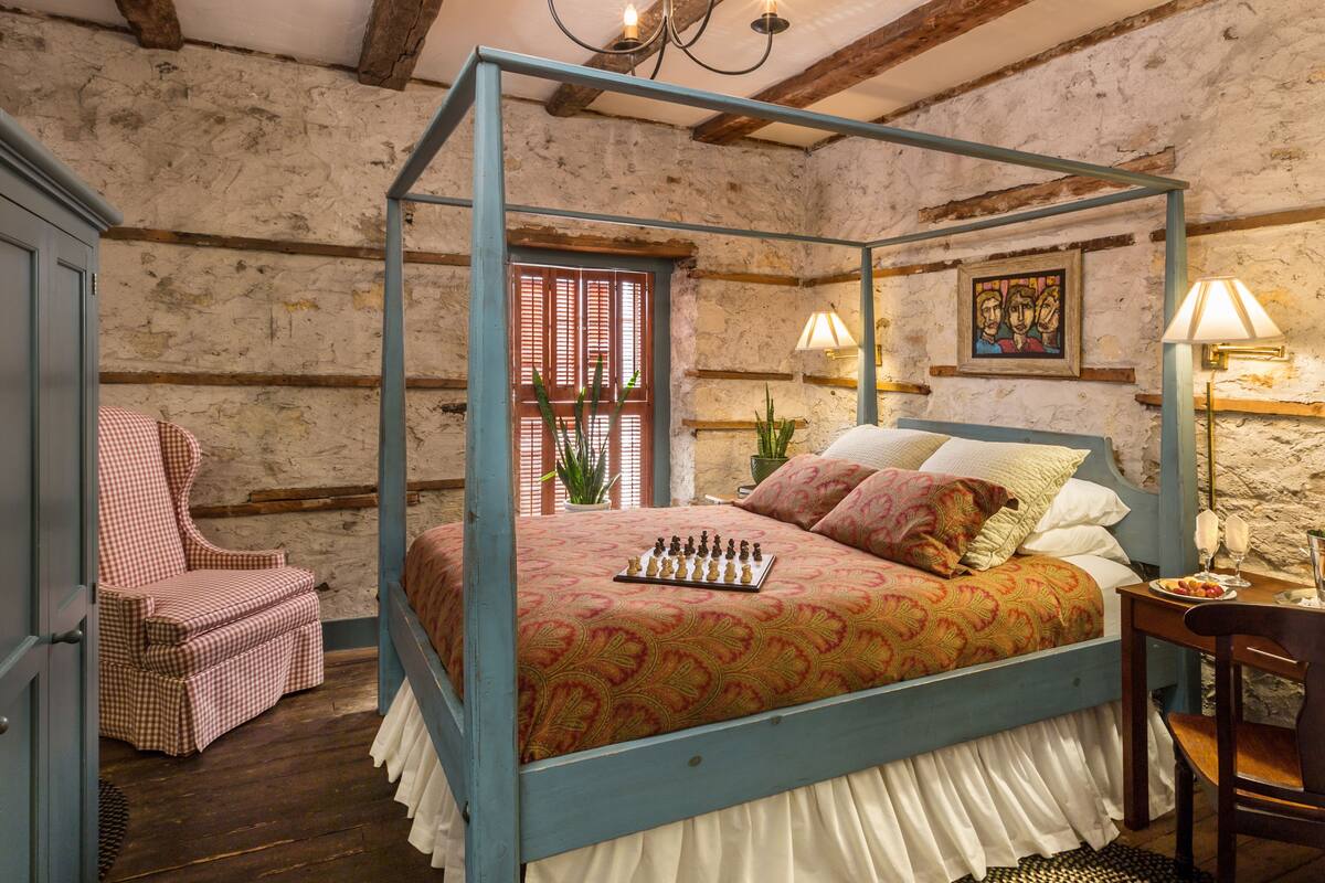 Uma cama com dossel, um tabuleiro de xadrez, uma poltrona xadrez e uma mesa com um copo de vinho.