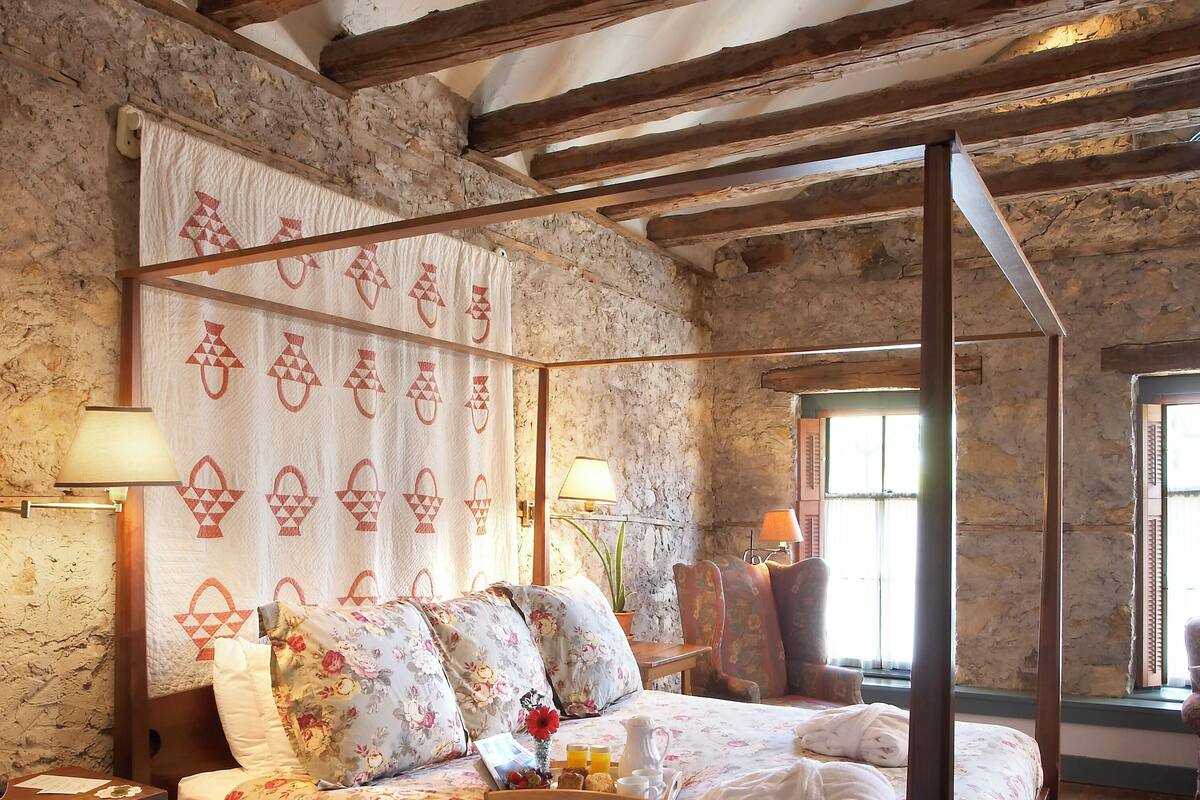 Uma cama com dossel com roupa de cama floral, uma mesa de cabeceira, um abajur e vista para as paredes de pedra e vigas de madeira do quarto.