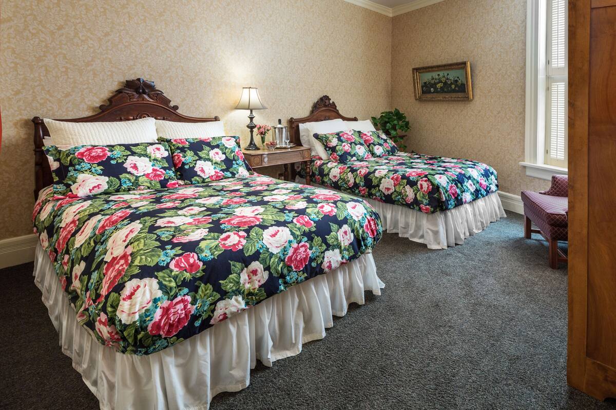 Quarto com duas camas, cada uma com colcha florida e saia branca franjada. Há uma mesa de cabeceira com abajur e um quadro pendurado na parede.