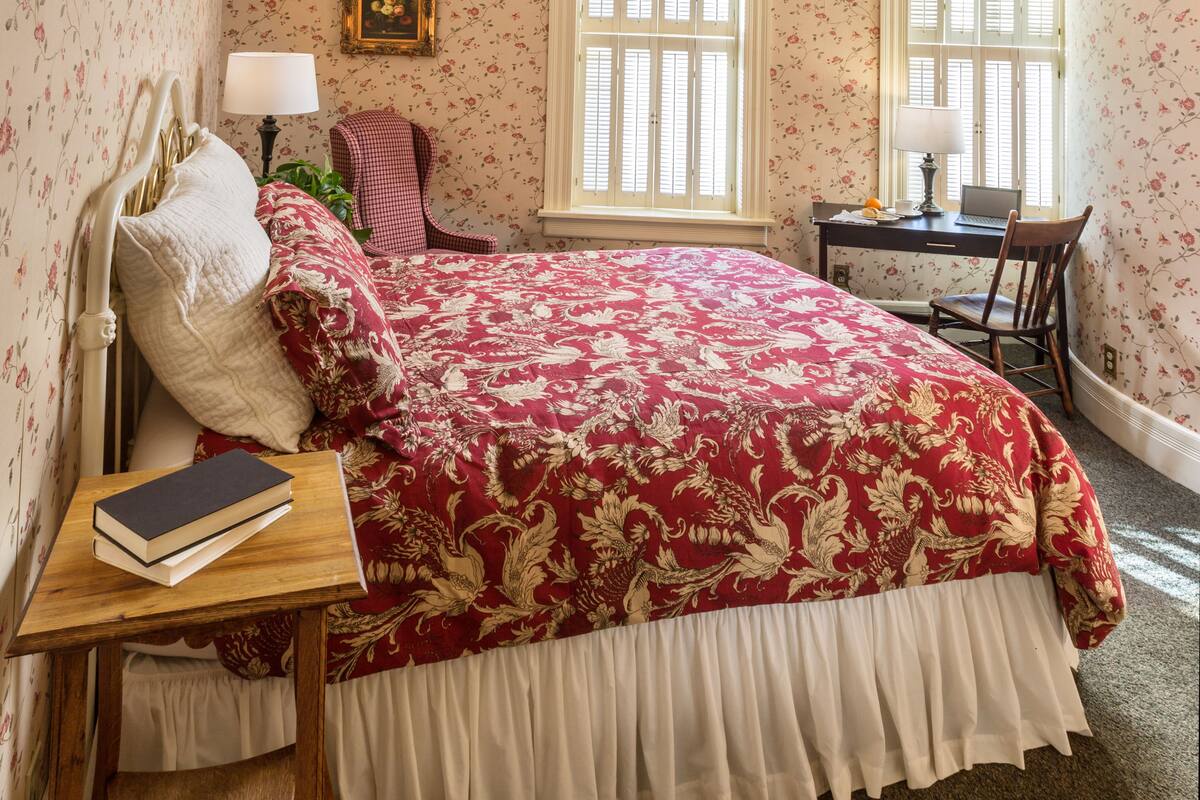 Quarto com papel de parede floral, colcha vermelha e branca, mesa de cabeceira de madeira com um livro, uma cadeira e uma escrivaninha com abajur.