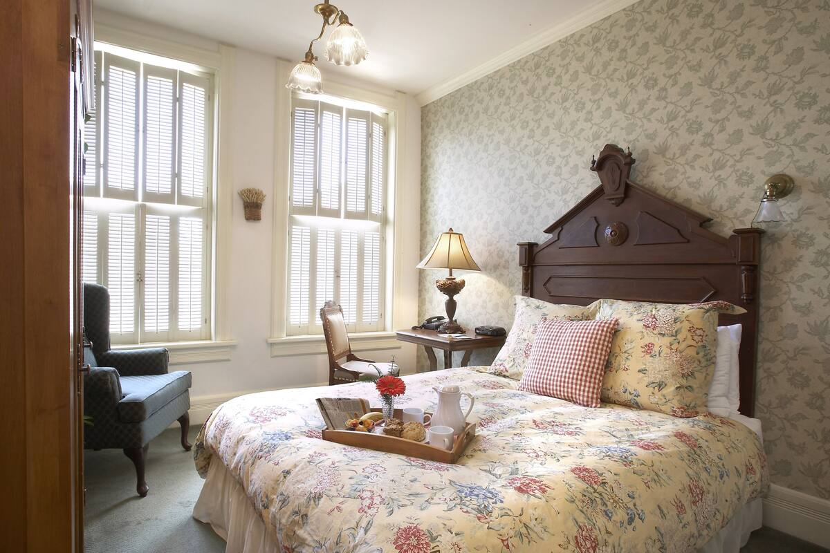 Quarto com colcha floral, cabeceira de madeira, mesa de cabeceira com abajur e uma cadeira.