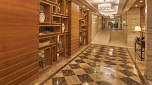 Um hall de hotel com piso de mármore quadriculado, estantes de madeira com objetos decorativos, um lustre e uma placa indicando os sanitários.