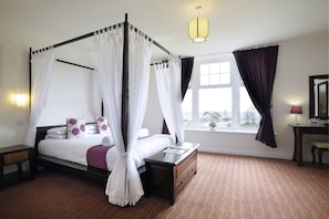 Uma cama com dossel e cortina, roupa de cama branca e roxa, mesa de cabeceira, abajur e vista do exterior pela janela.