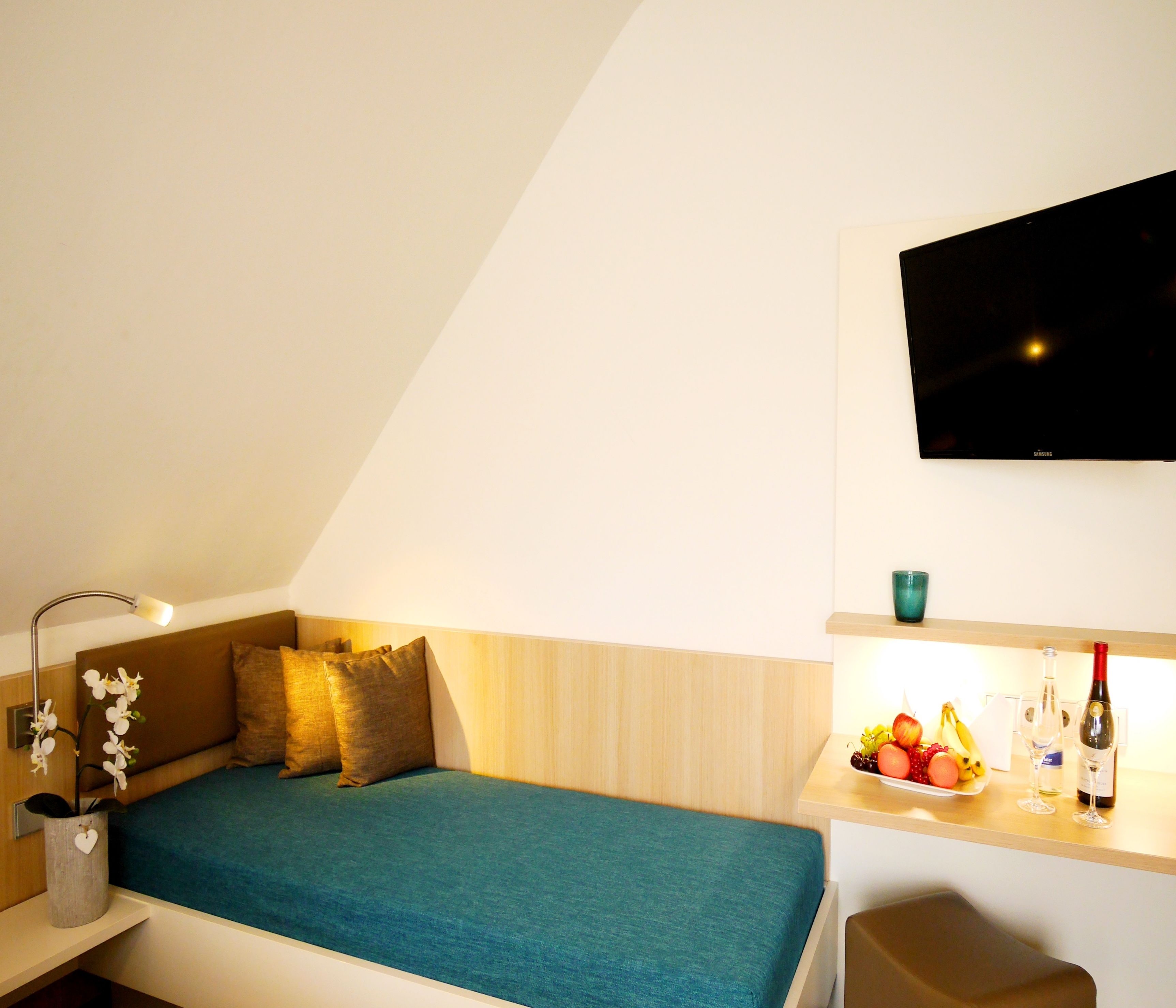 Quarto com cama, mesa de cabeceira com abajur, TV na parede e uma pequena estante com frutas e uma garrafa.