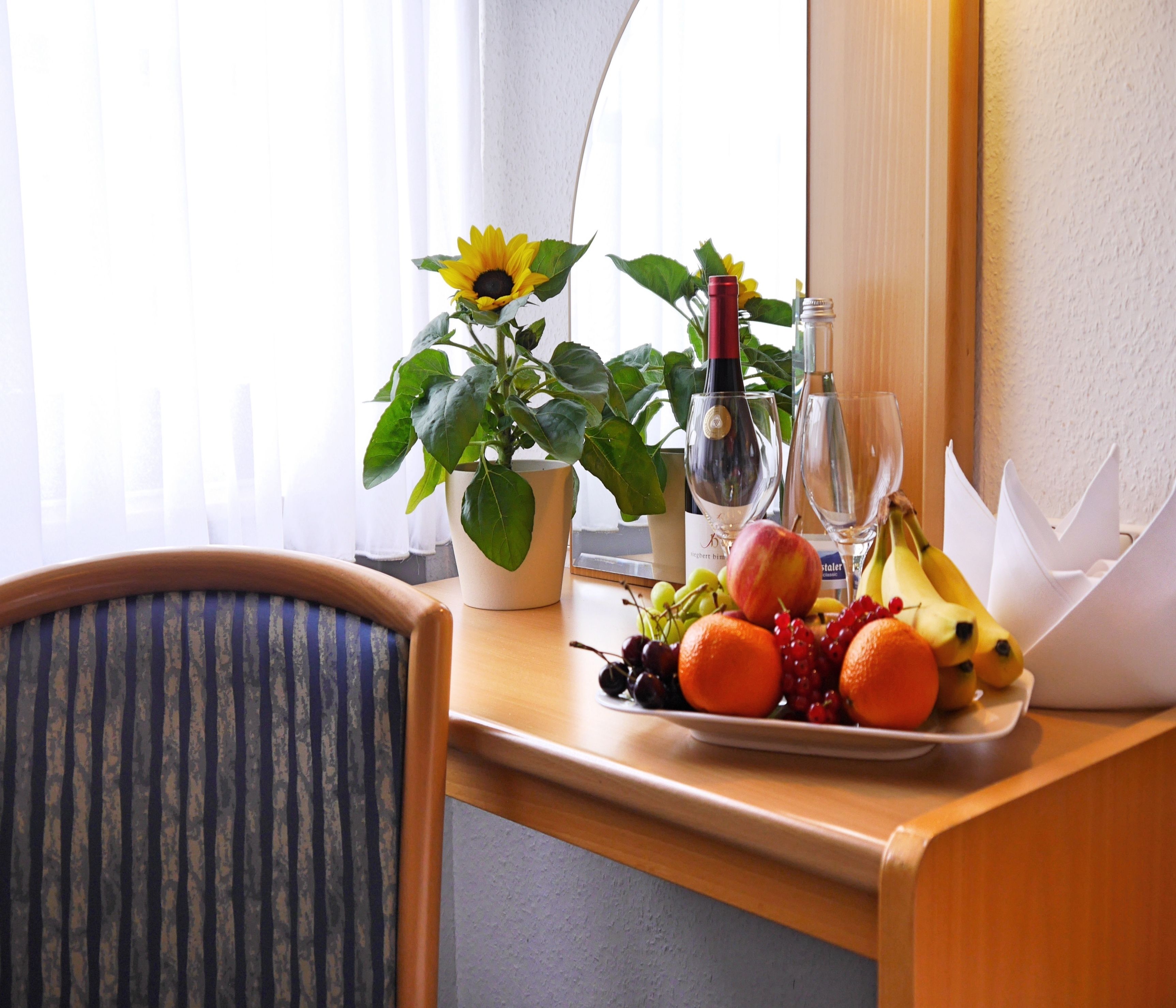 Quarto de hotel com escrivaninha de madeira, planta em vaso, garrafa de vinho, taças de vinho e bandeja de frutas.
