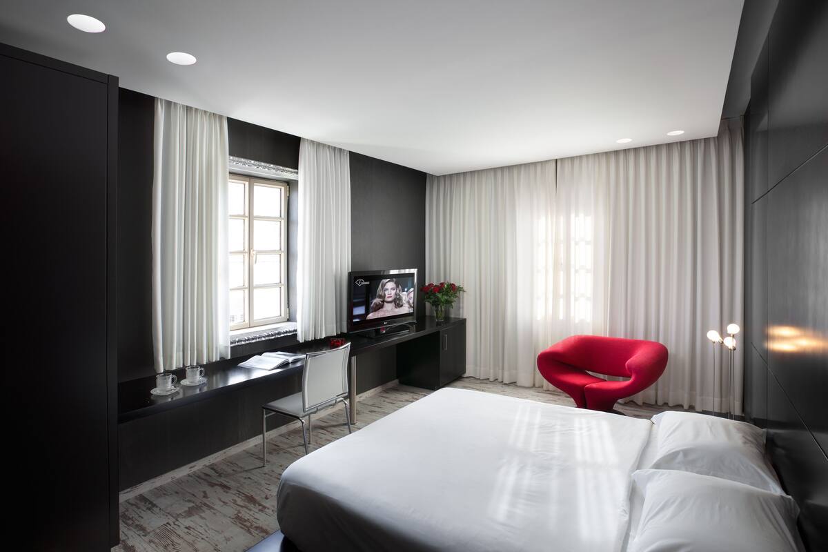 Quarto de hotel moderno com uma cama grande, uma mesa de escritório com um computador, uma cadeira vermelha e uma televisão.