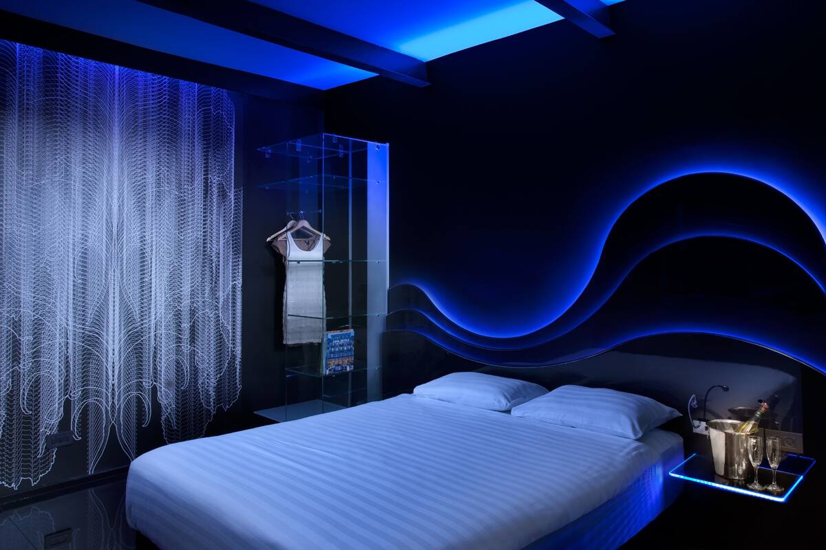 Quarto moderno com divisória de vidro, cama e mesa de cabeceira com garrafa e copos.