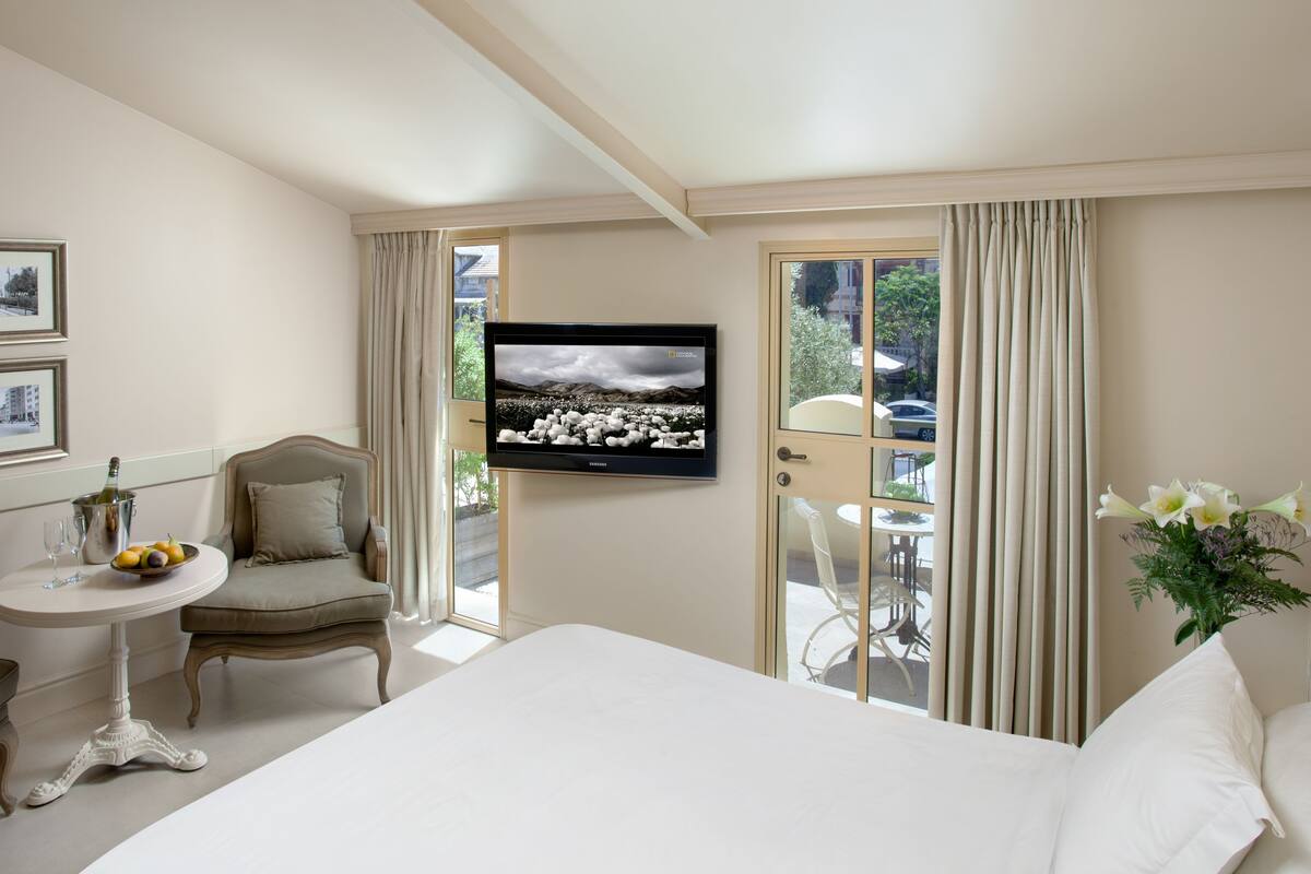 Quarto de hotel com cama, televisão, uma pequena mesa com garrafa e copos, uma cadeira e janela com cortinas.