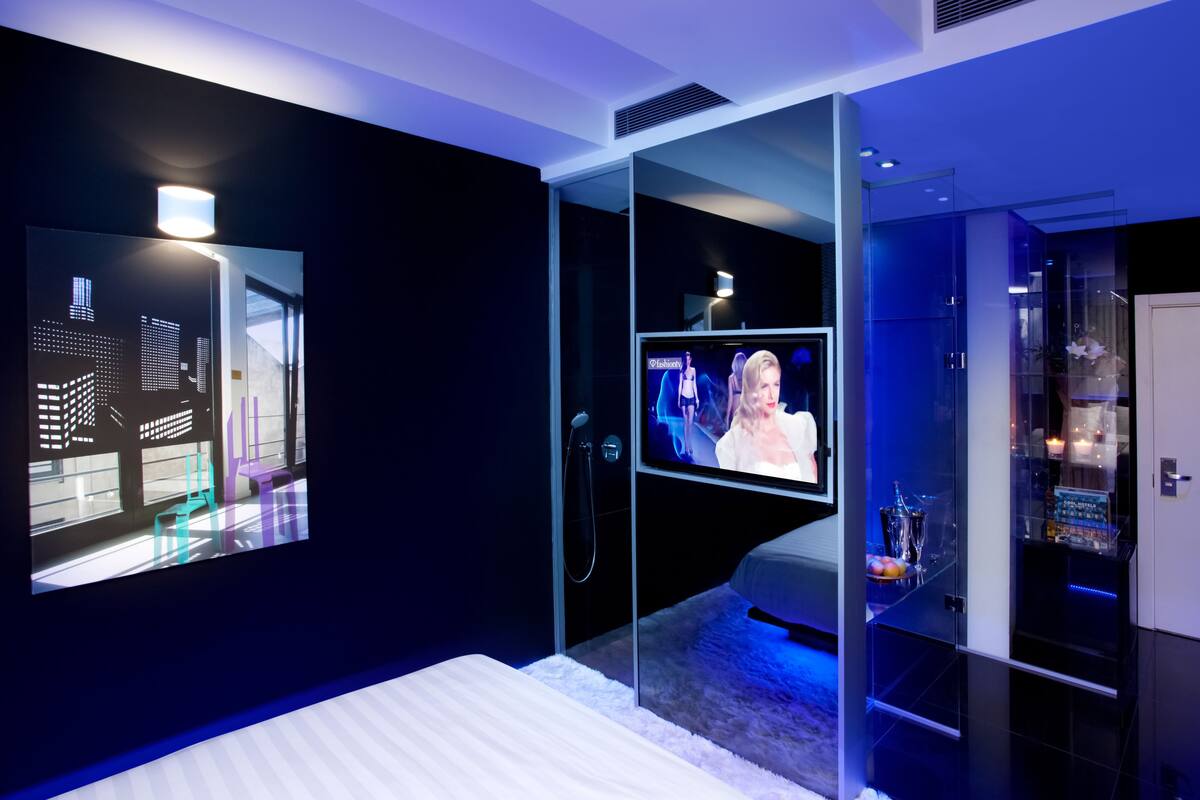 Quarto de hotel moderno com uma televisão grande, chuveiro com box de vidro e minibar.