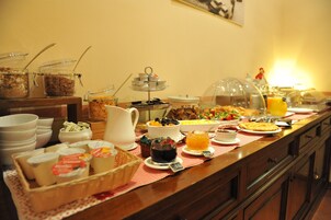 Buffet com diversos alimentos e bebidas, incluindo estação de café, frutas, pastéis e dispensador de suco.