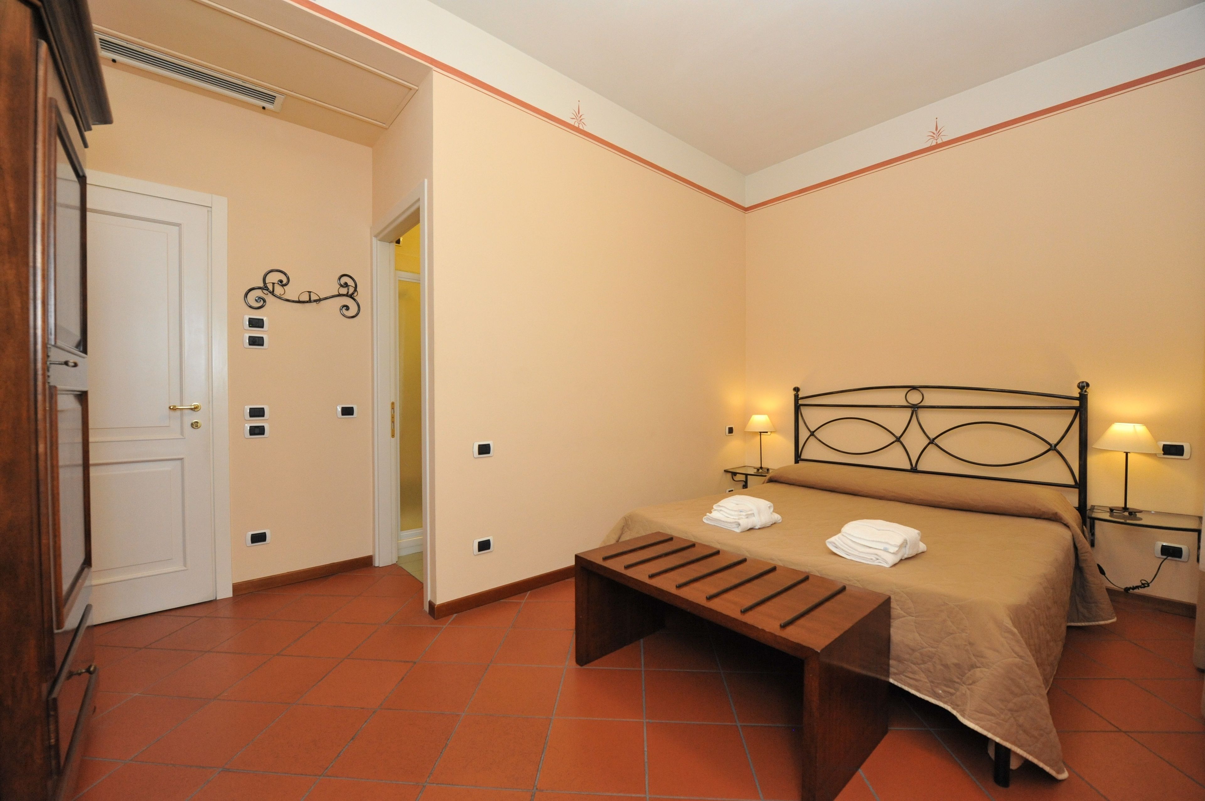 Quarto de hotel com cama, luminárias de cabeceira, um banco e uma porta.