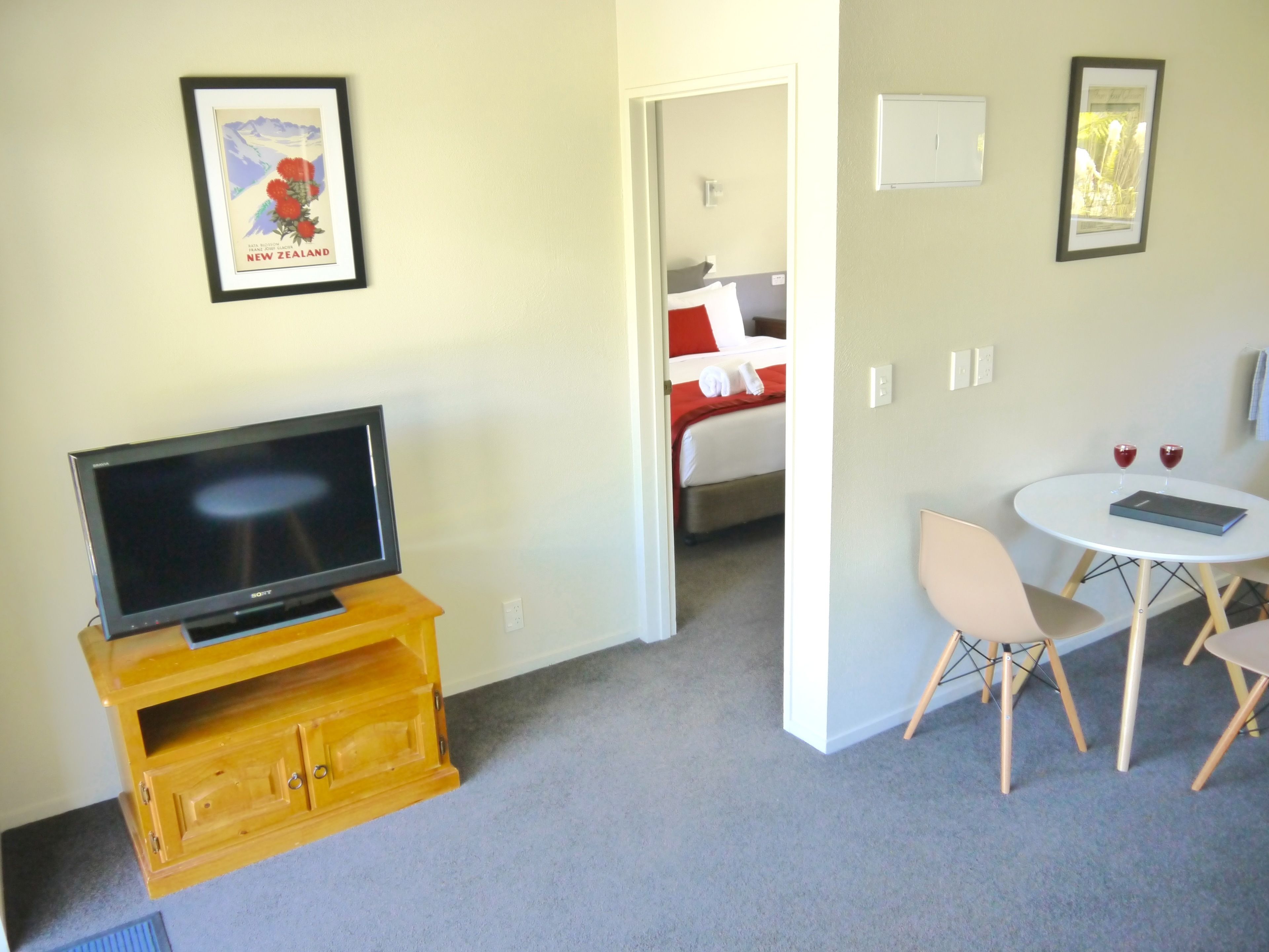Quarto com televisão sobre suporte de madeira, quadro emoldurado da Nova Zelândia, pequena mesa com duas cadeiras e cama com cobertor vermelho ao fundo.