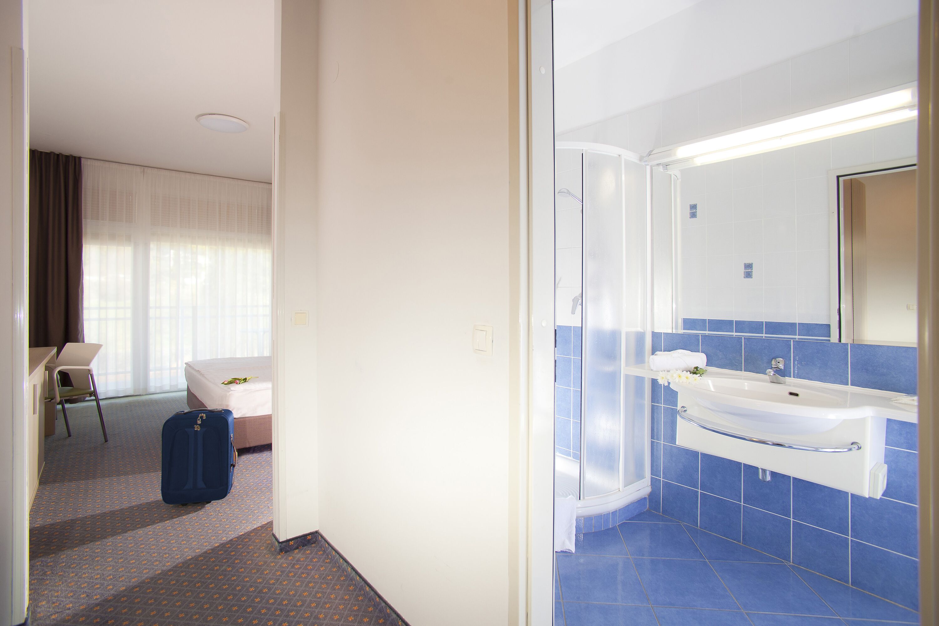Quarto de hotel com cama, banheiro com duas pias e mala no chão.