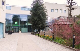 Um hotel Holiday Inn com fachada de vidro moderna, área de estar ao ar livre e um jardim bem cuidado com flores e uma grande árvore.