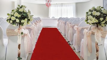 Capela de hotel com cadeiras brancas, tapete vermelho e decoração floral.