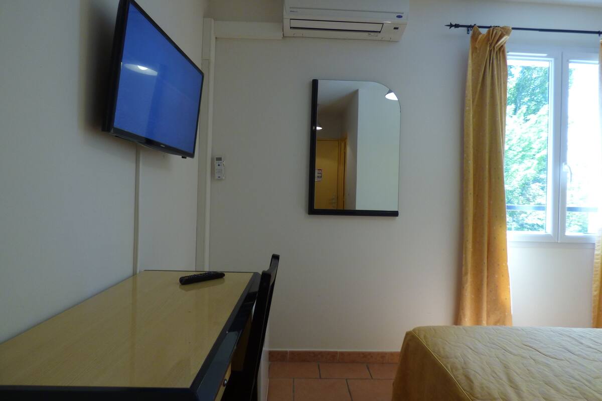 Quarto com cama, escrivaninha, televisão, ar condicionado, espelho e janela com cortinas.
