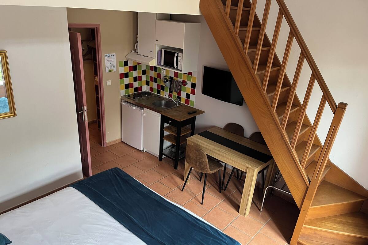 Um quarto com cama, escada de madeira, cozinha com micro-ondas e uma pequena mesa.