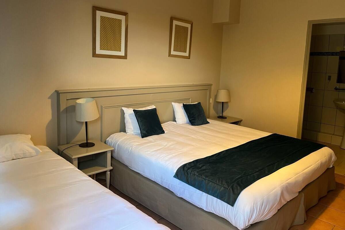 Quarto de hotel com duas camas, cada uma com roupa de cama branca e almofadas escuras. Há duas mesas de cabeceira com abajures, e o quarto apresenta uma paleta de cores neutras.