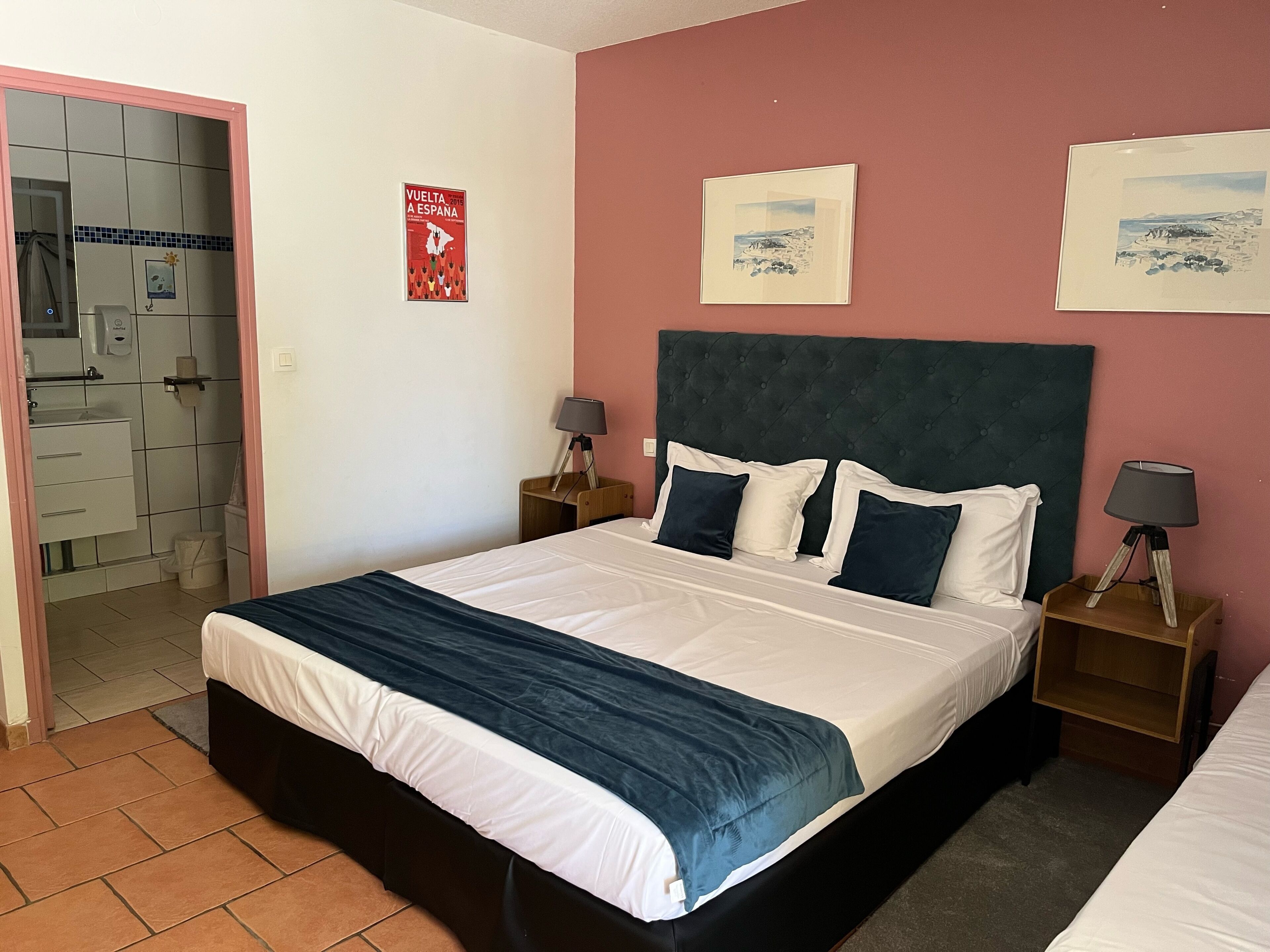 Quarto com cama, duas mesas de cabeceira, um criado-mudo, um cartaz na parede e um banheiro visível por uma porta aberta.