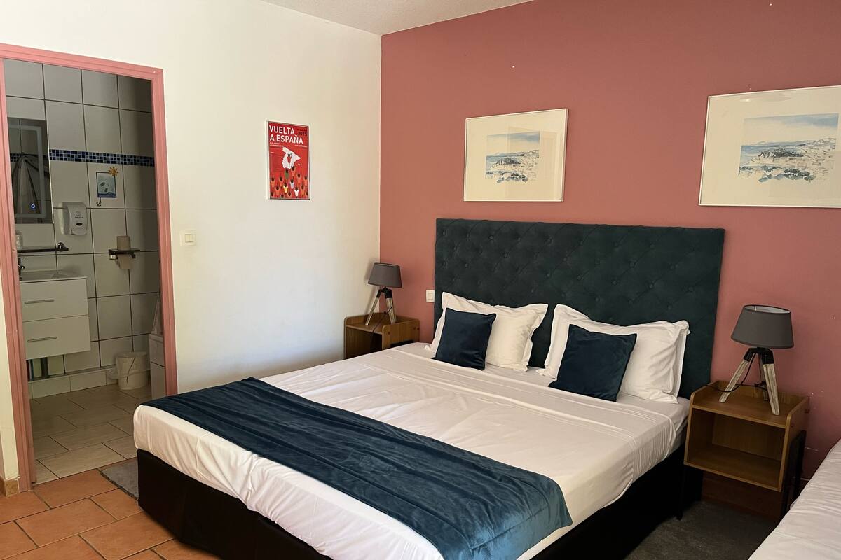 Quarto com cama, duas mesas de cabeceira, um criado-mudo, um cartaz na parede e um banheiro visível por uma porta aberta.