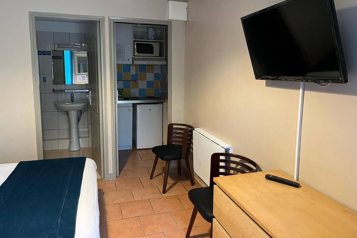 Quarto compacto com cama, escrivaninha, televisão, cadeira, pequeno espaço de cozinha e banheiro.
