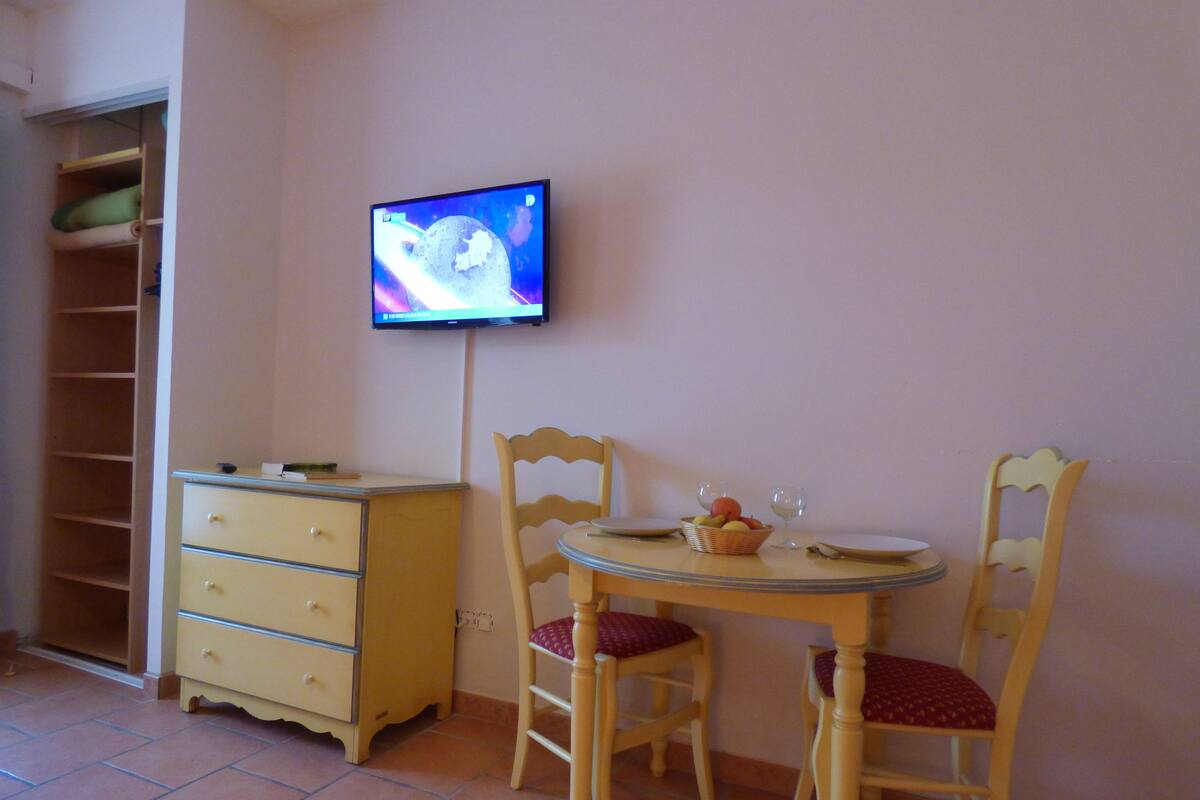 Uma sala de jantar com uma mesa, cadeiras, uma televisão fixada na parede e um armário de madeira.