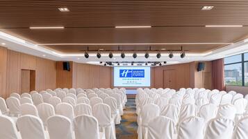 Uma sala de conferências com fileiras de cadeiras brancas, uma tela grande e teto de madeira.