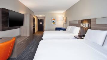 Quarto de hotel com duas camas, uma escrivaninha, uma cadeira laranja, uma televisão de tela plana e obras de arte nas paredes.