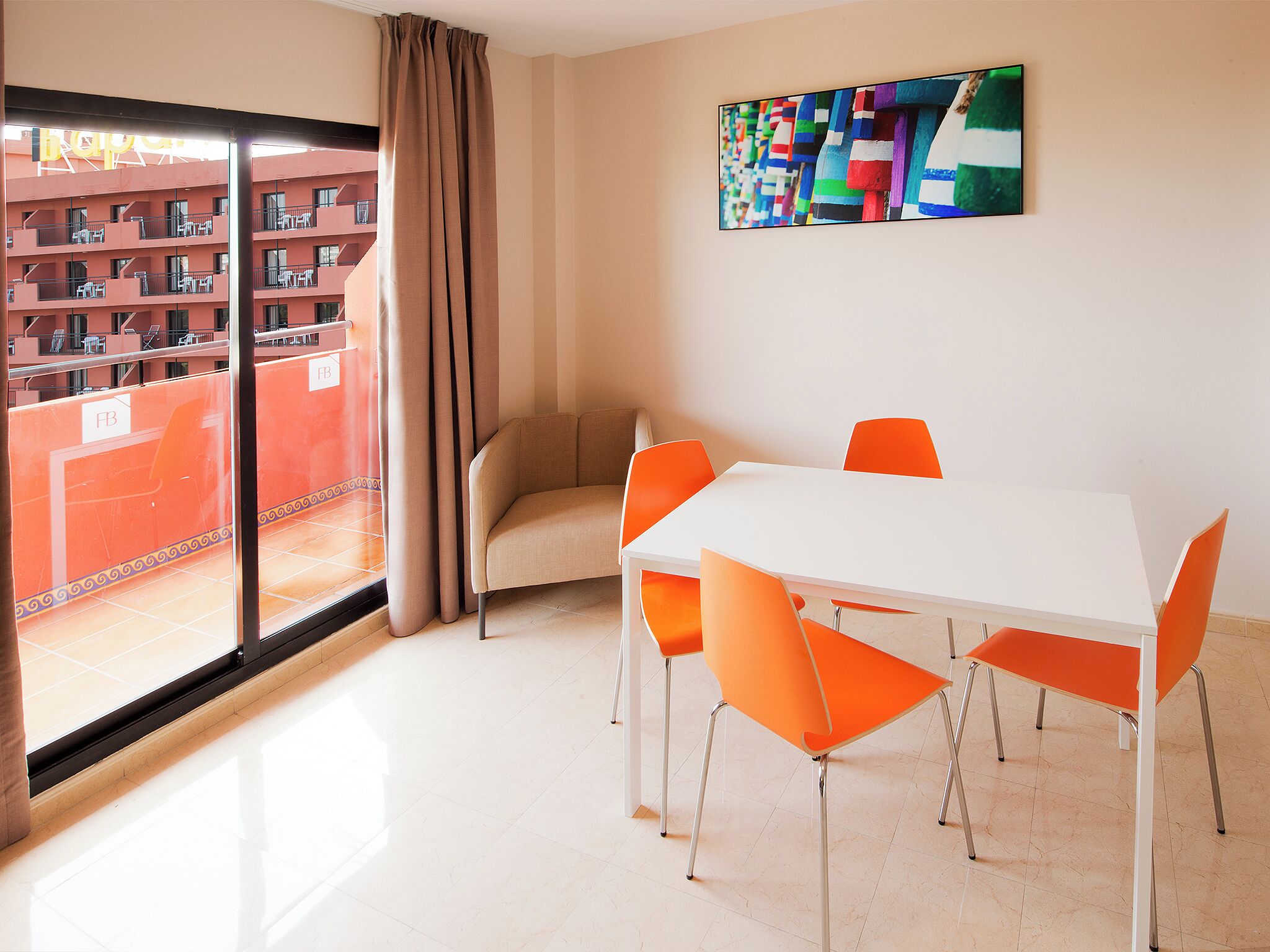 Quarto moderno com uma mesa branca e cadeiras laranja, uma janela ampla com vista para um edifício e uma pintura abstrata colorida na parede.