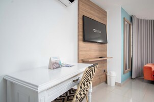 Quarto de hotel moderno com escrivaninha branca, televisão montada, parede de destaque em madeira e janela com cortinas.