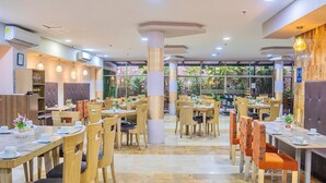 Um restaurante espaçoso com várias mesas e cadeiras, grandes janelas com vista para o verde lá fora, e um design moderno no teto com iluminação embutida.