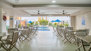 Amplo espaço interno com várias mesas e cadeiras dispostas para refeições, uma piscina no centro e ventiladores de teto.