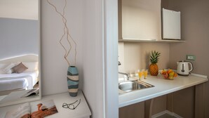 Quarto de hotel com uma cama, mesa de cabeceira com um vaso e copos, cozinha com pia, geladeira, tigela de frutas e chaleira.