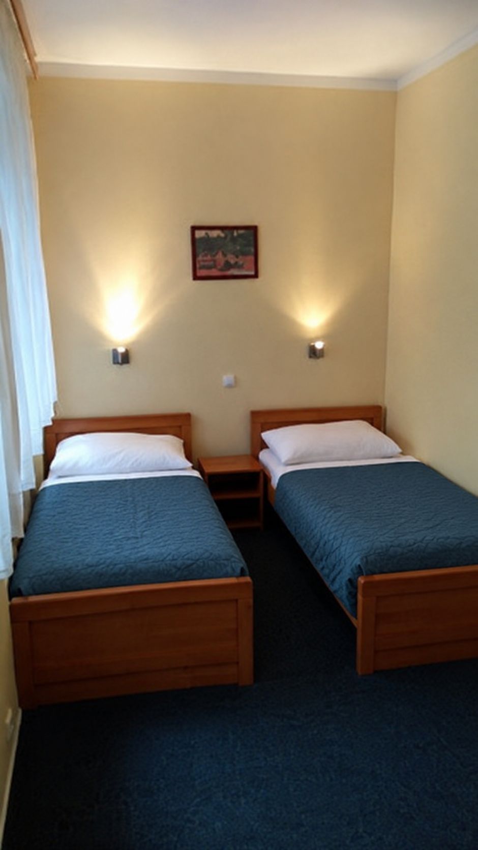 Quarto com duas camas de madeira, cada uma com colcha azul e travesseiros brancos, e duas luminárias fixadas na parede.