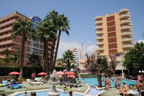 Área de piscina de hotel com vários níveis de edifícios, palmeiras e uma fonte.