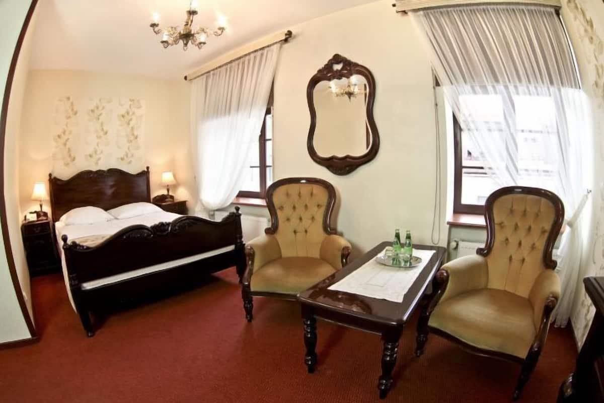 Quarto de hotel com cama de madeira, duas poltronas, uma mesinha, um espelho e um candelabro.