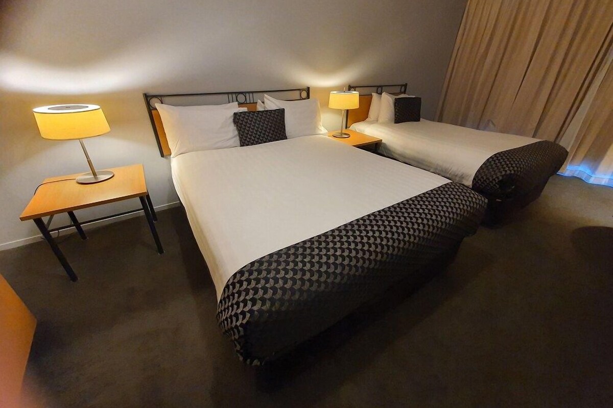 Quarto de hotel com duas camas, uma mesa de cabeceira, uma luminária e uma janela com cortinas.
