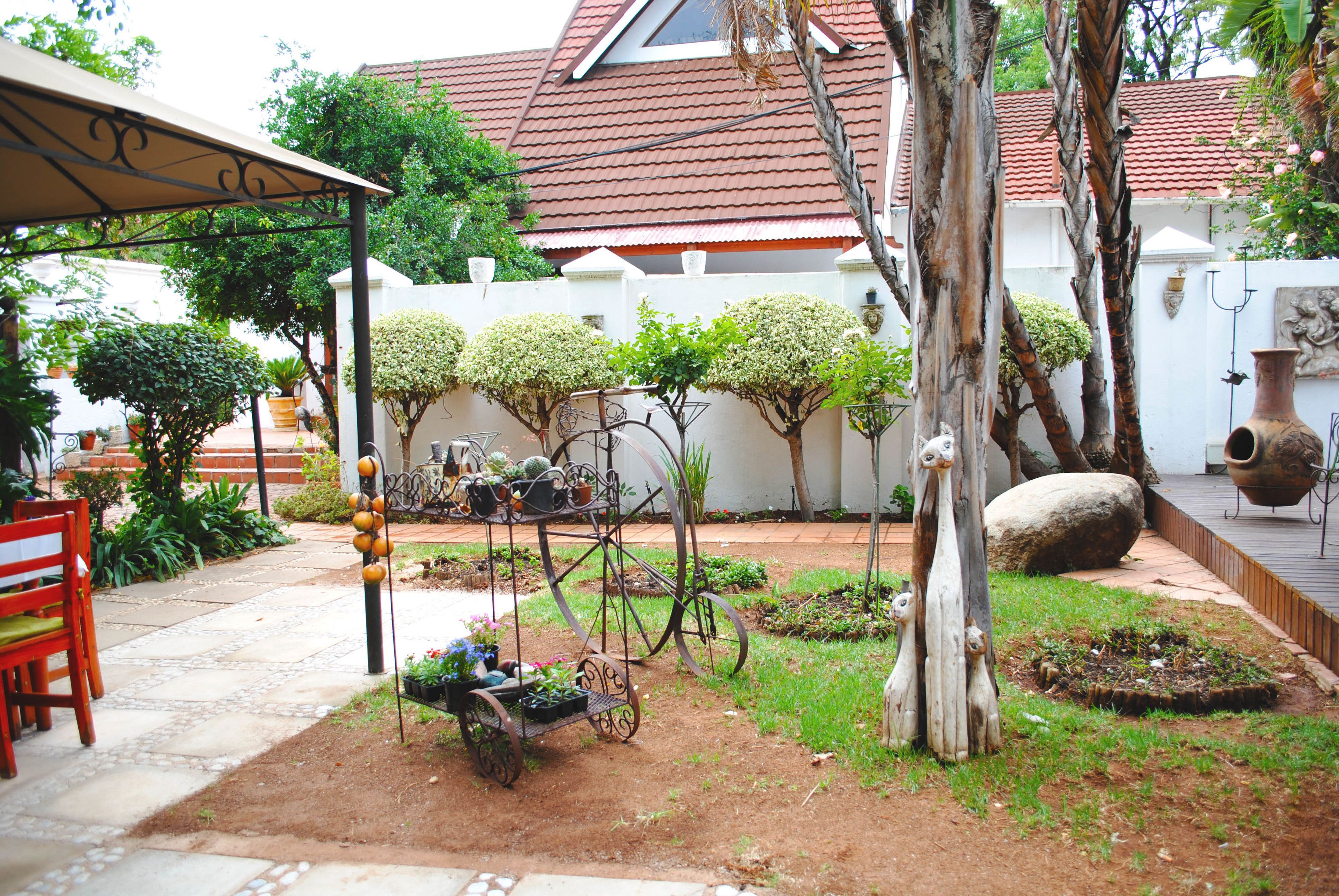 Um jardim com um banco de madeira, um carrinho de metal com plantas e uma parede branca com telhado vermelho.