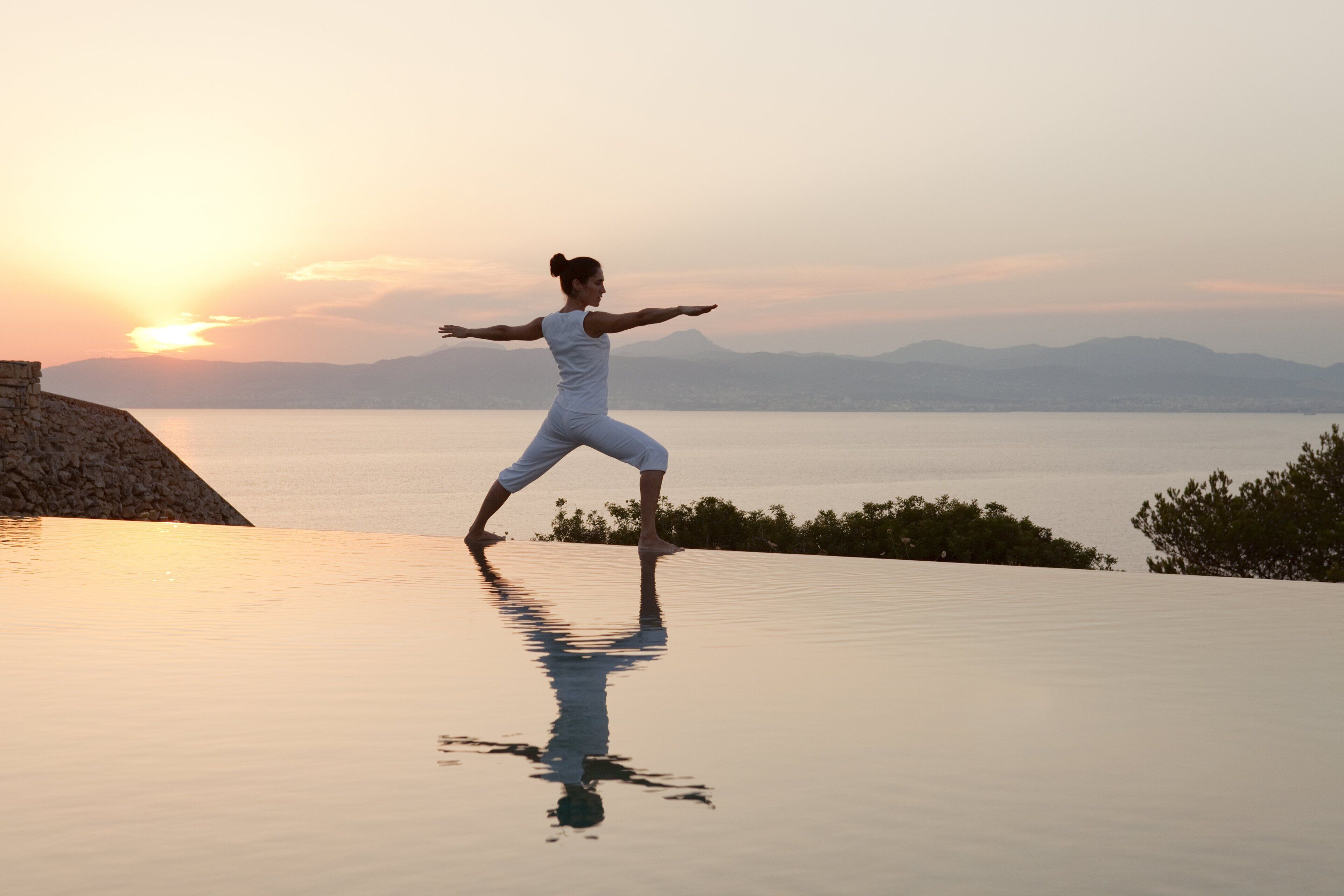 Uma pessoa fazendo uma postura de yoga em uma piscina refletiva com um pôr do sol e montanhas ao fundo.