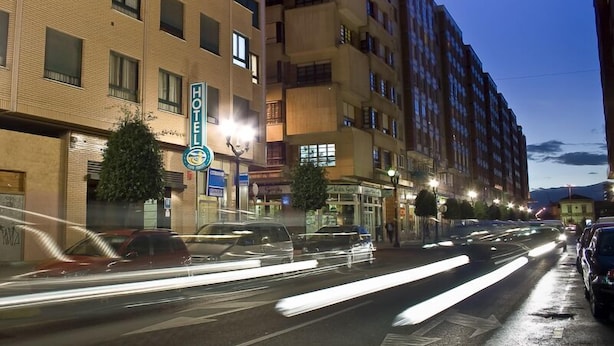 Um hotel com vários andares, um letreiro chamativo com a palavra 'HOTEL', uma entrada visível e vista para a rua com carros e uma faixa de pedestres.