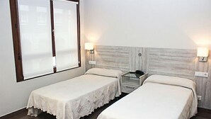 Quarto de hotel com duas camas de solteiro, uma pequena mesa, duas luminárias fixadas na parede e uma janela com persianas.