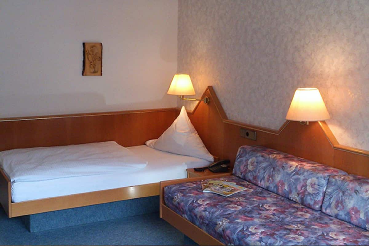 Quarto de hotel com cama, sofá, duas luminárias fixadas na parede e uma mesa pequena com um controle remoto.