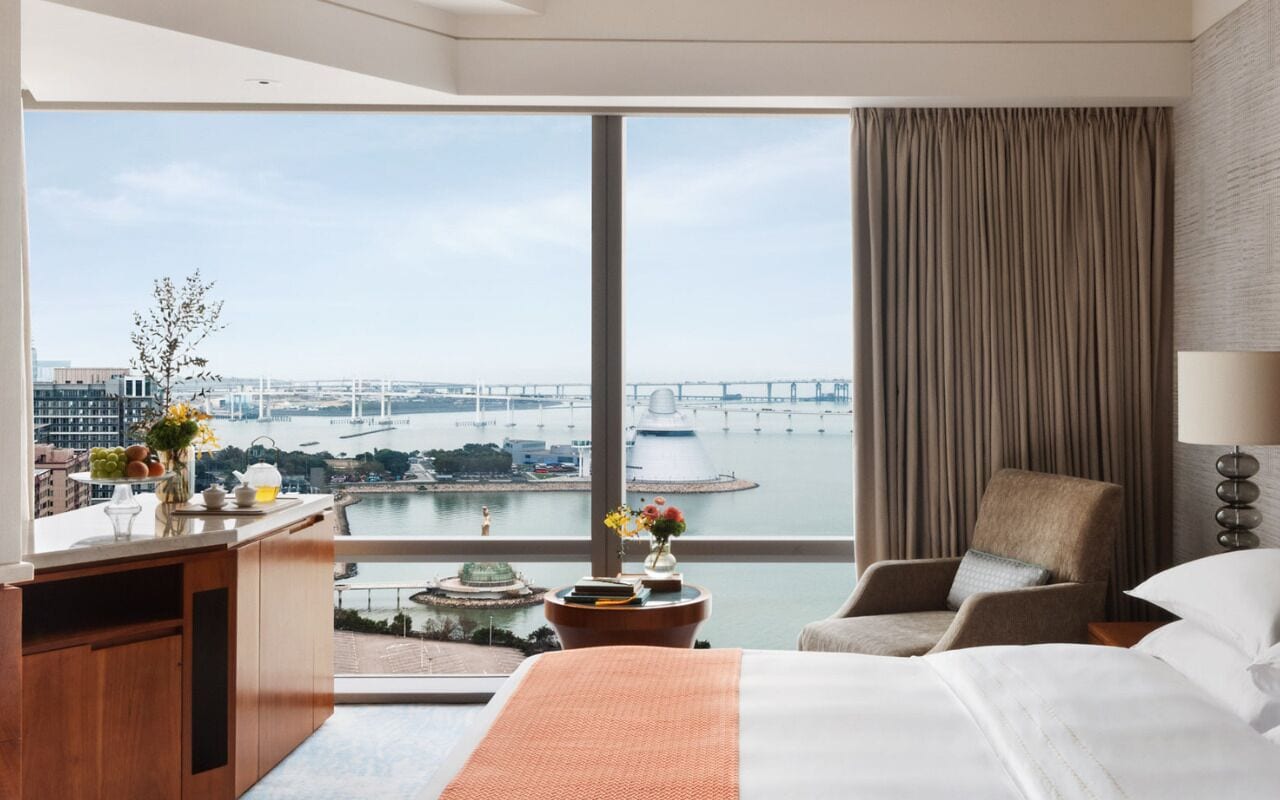 Quarto de hotel com uma grande janela, cama, escrivaninha, cadeira e vista para uma ponte e para a água.