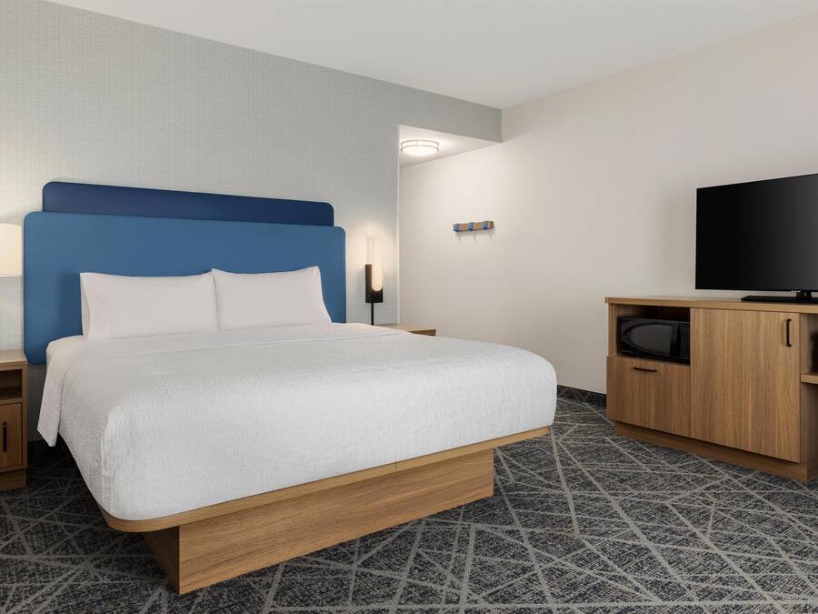 Quarto de hotel com cama, televisão, máquina de café e mesa de cabeceira.