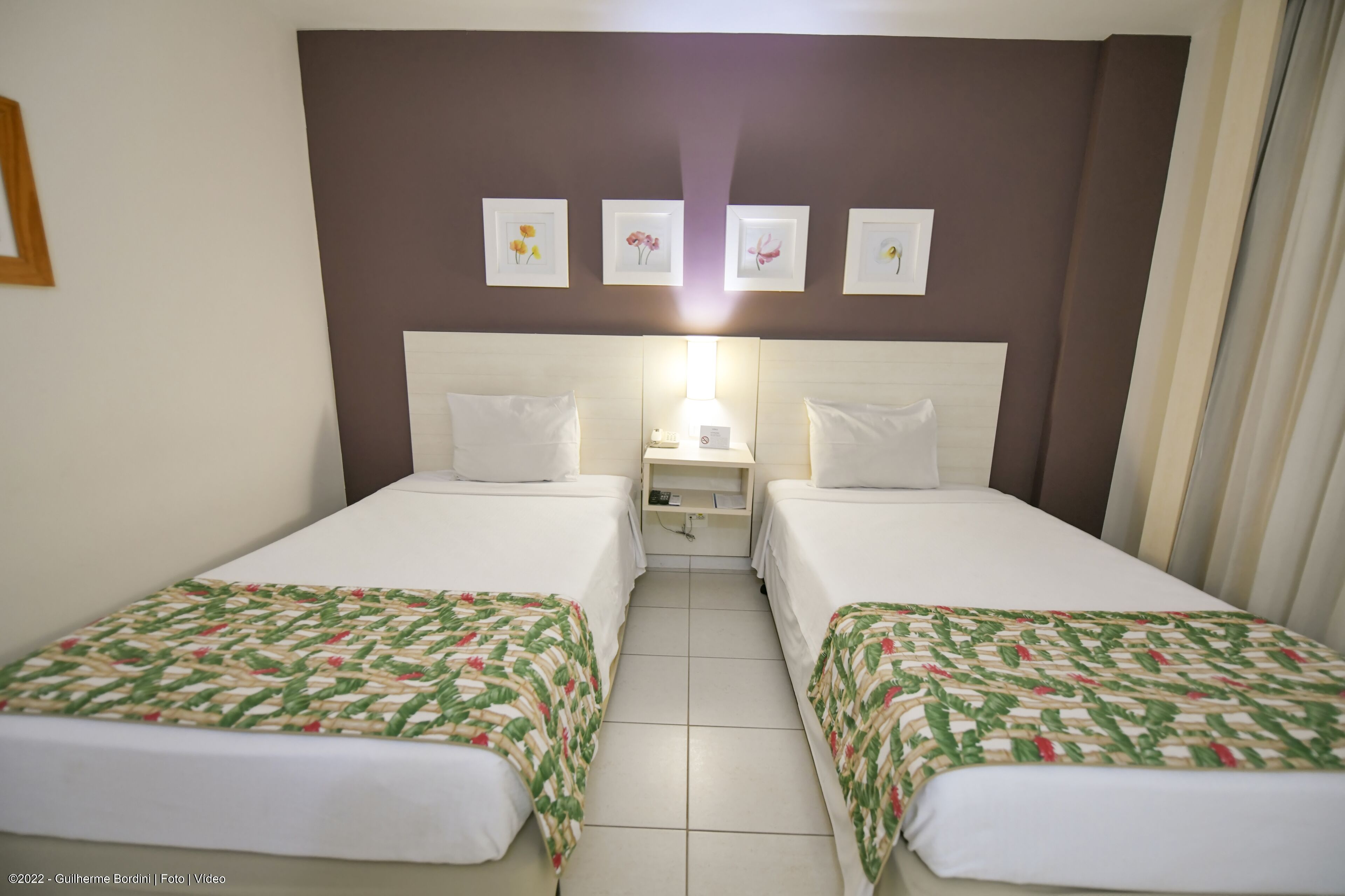 Quarto de hotel com duas camas, uma mesinha de cabeceira e uma luminária fixada na parede.