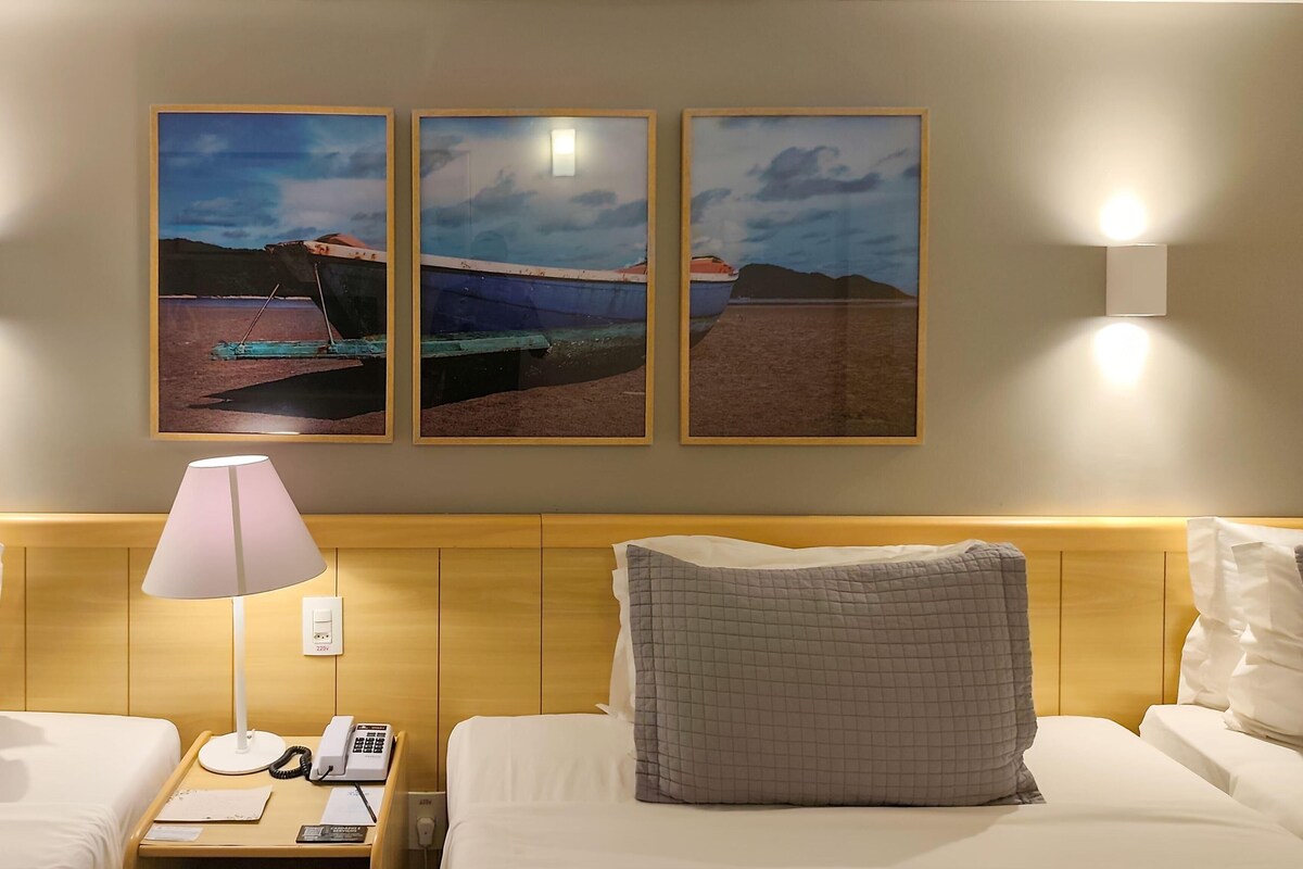 Quarto de hotel com cama, abajur, telefone e uma obra de arte na parede retratando uma cena de praia.